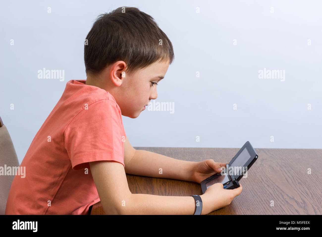 Young boy playing handheld video game at a table. Against grey ...