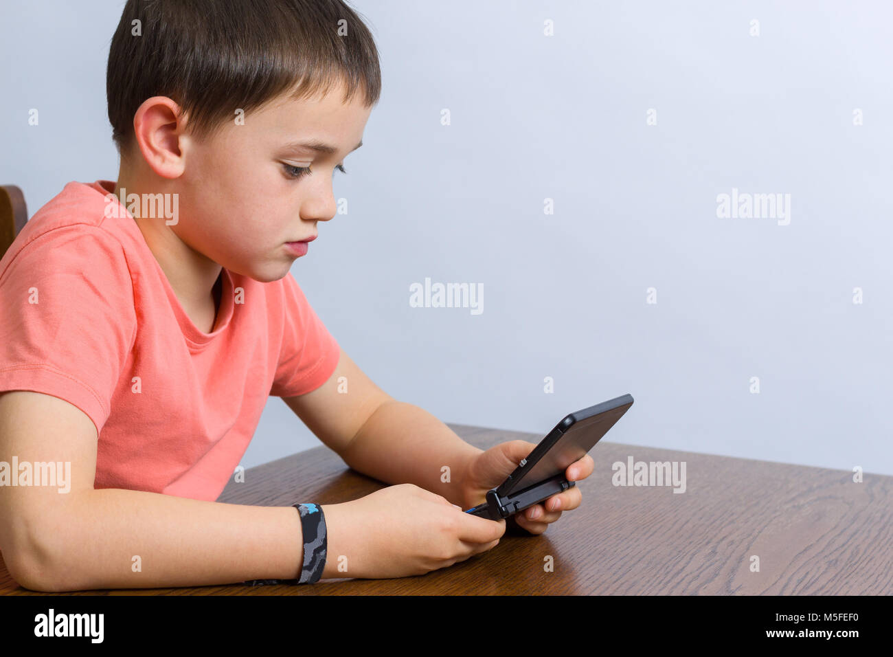 Young boy playing handheld video game at a table. Against grey ...