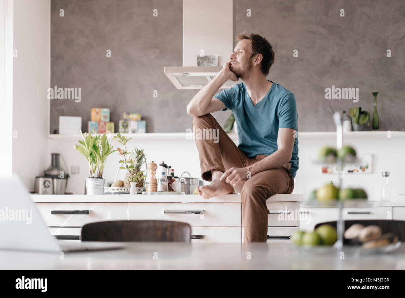 Smilingg man sitting on kitchen counter looking at distance Stock Photo ...