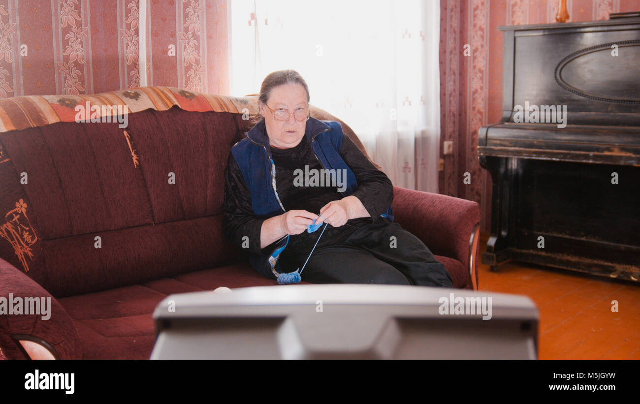 Old lady home at sofa - senior woman watching television and knits wool ...
