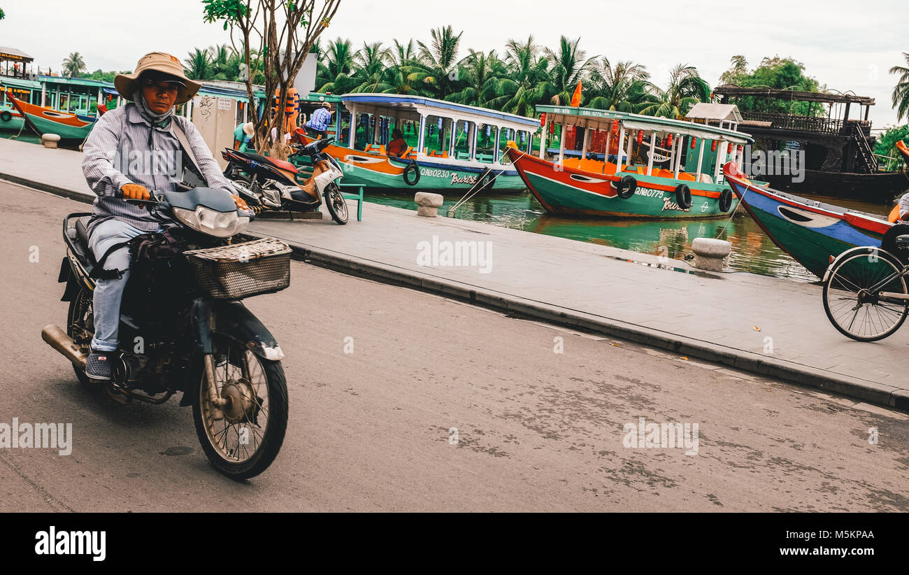 A woman on a moped drives past next to the river boats in Hoi An ...