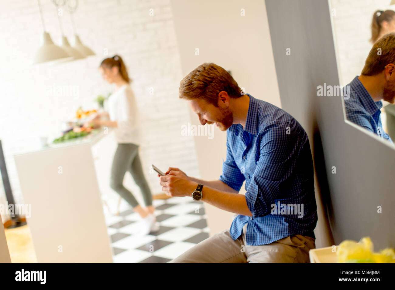 Young man uses a cell phone in modern kitchen while young woman cooks ...