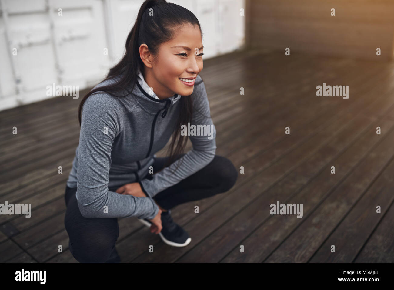 Smiling young Asian woman in exercise clothing crouching on the ground ...