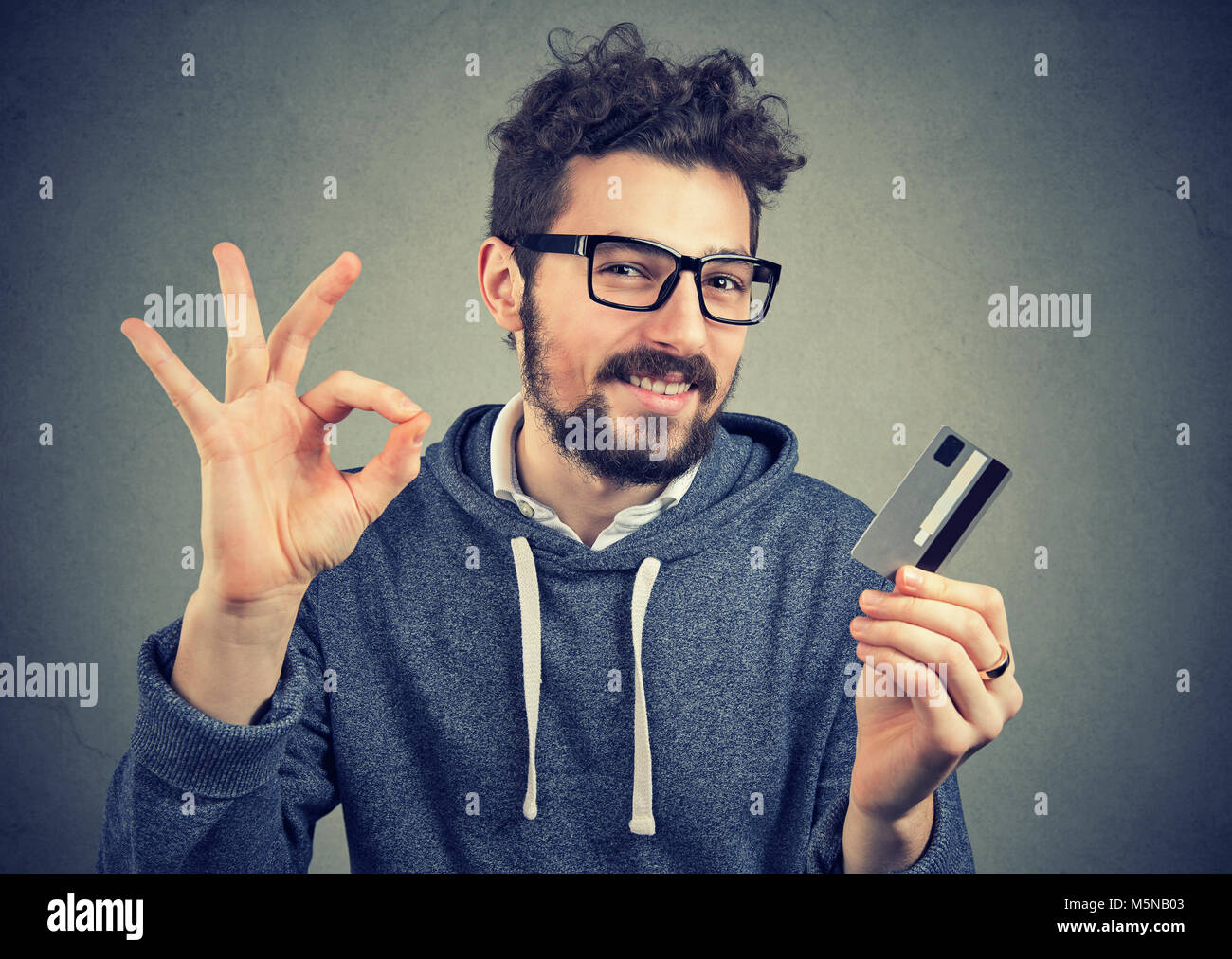 Young hipster man showing OK gesture and credit card smiling happily at ...