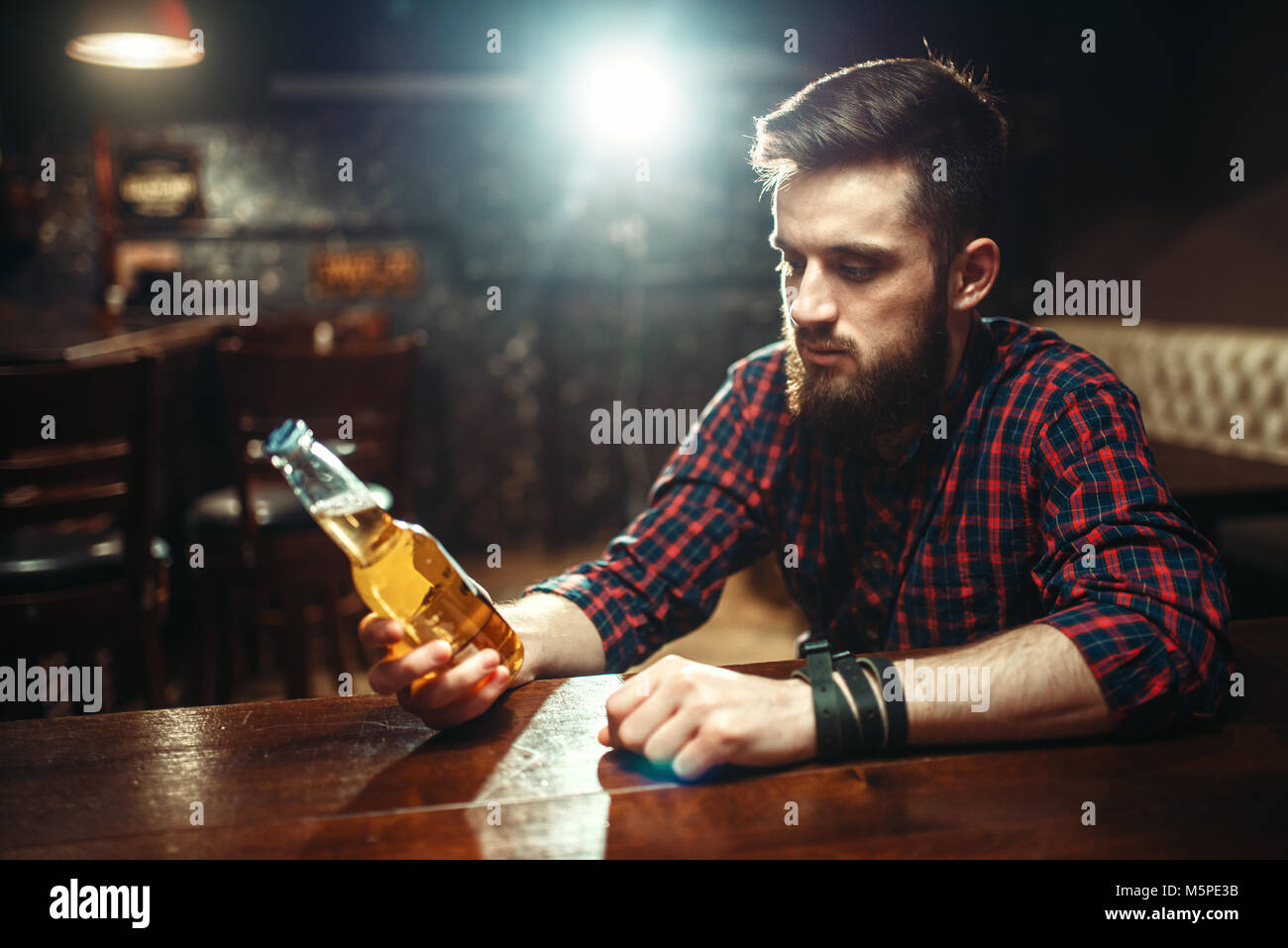 Sad man sitting at the bar counter and holds the bottle with alcohol ...