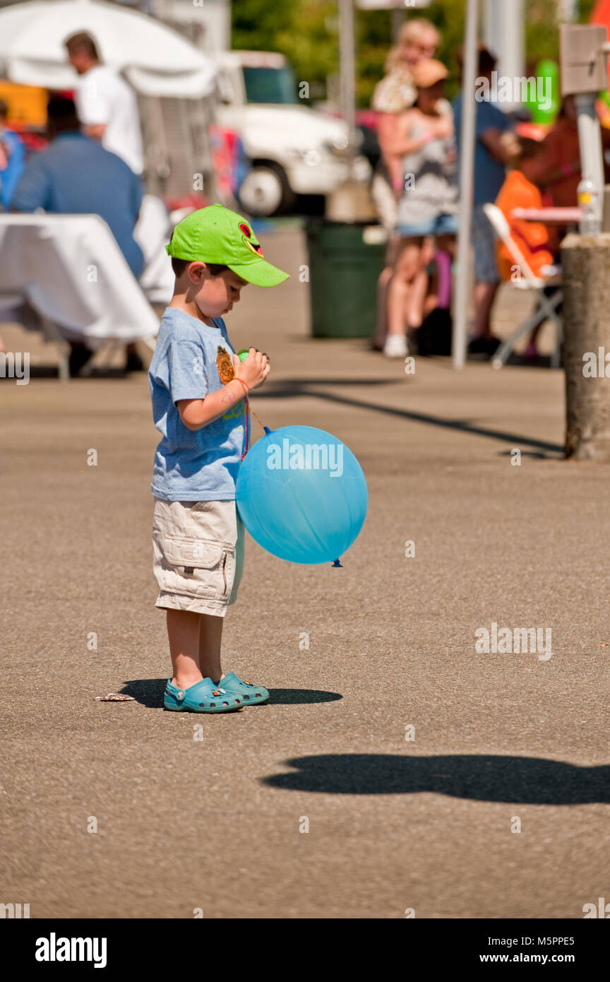 A boy with a green baseball cap tying a big blue balloon at a summer ...