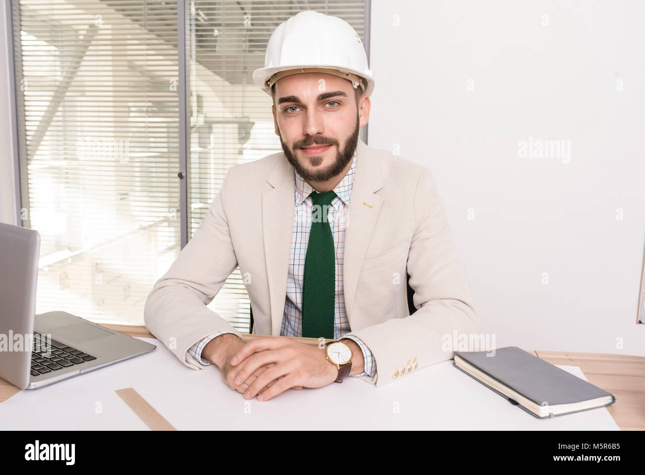 Portrait of handsome young engineer wearing white hardhat sitting at ...