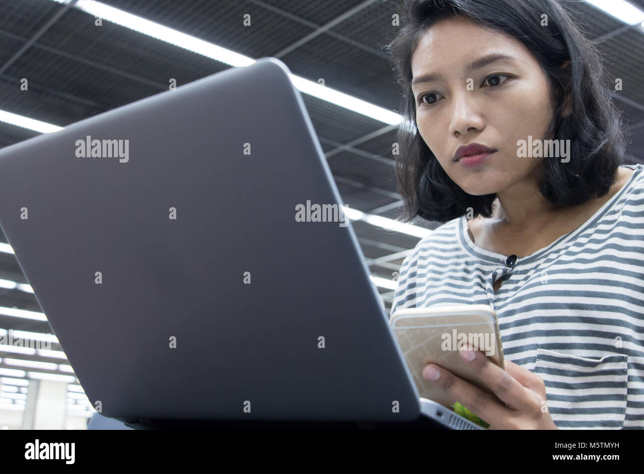 A young woman is working on a computer in an airport lobby while ...