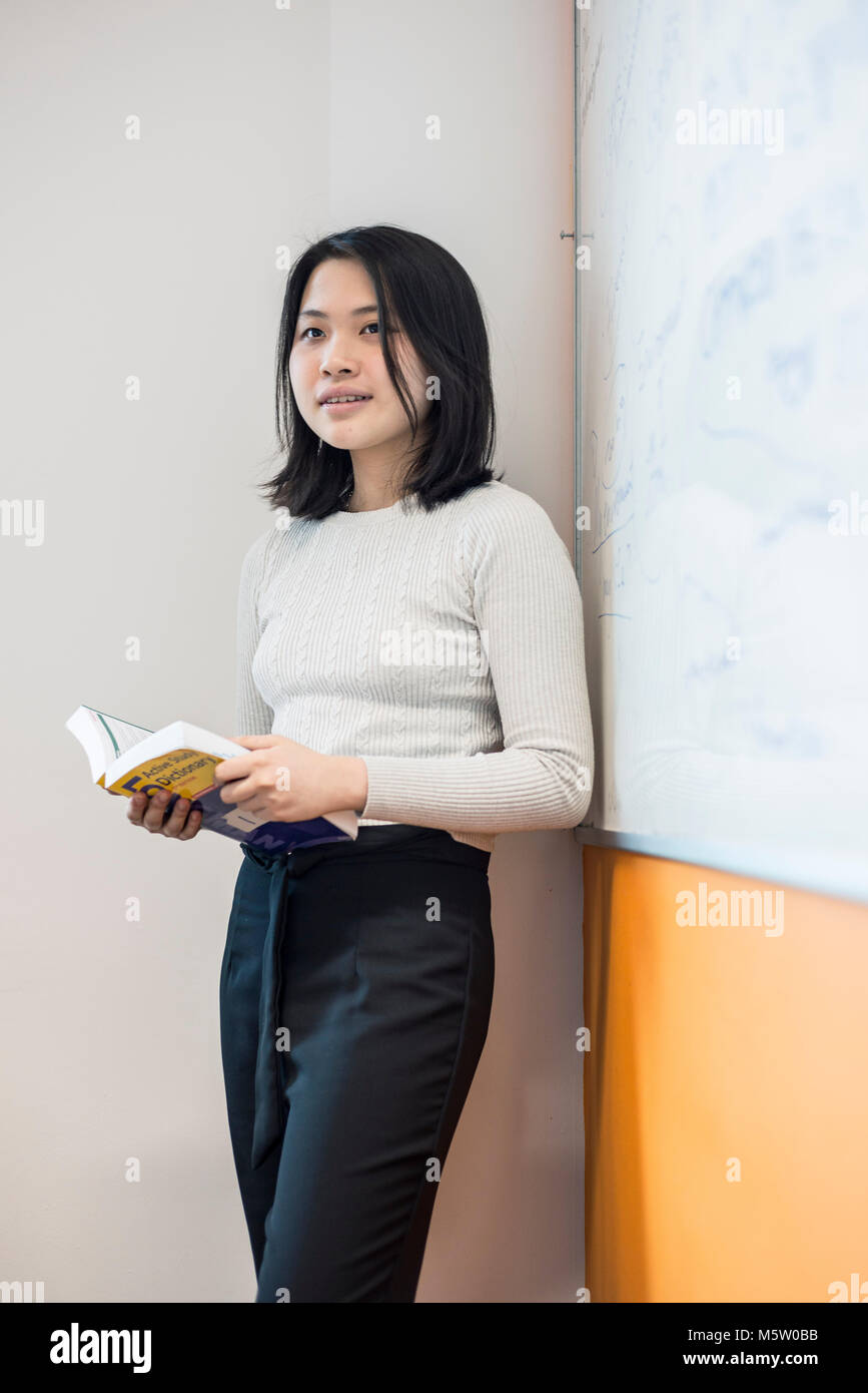 a young asian international student stands in a classroom with a book ...