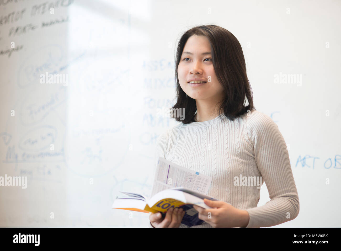 a young asian international student stands in a classroom with a book ...