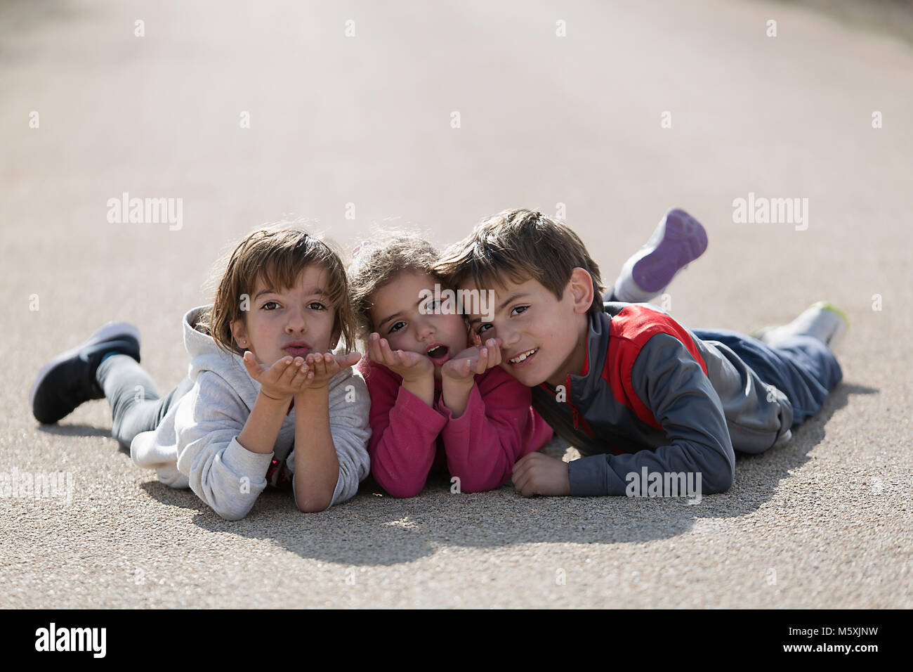 Three children lying on the ground of a road facing the camera ...