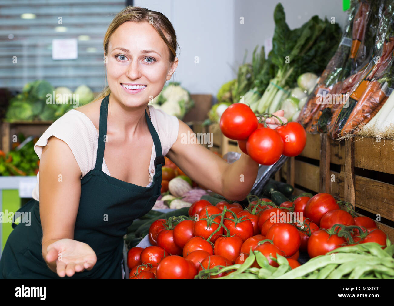 Portrait of young happy woman in apron selling organic tomatoes in shop ...