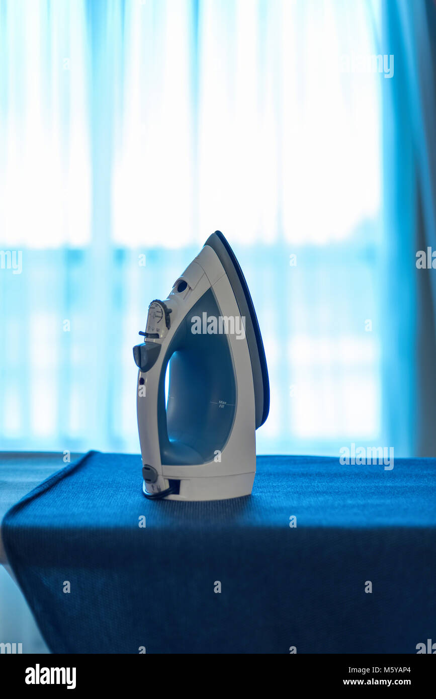 hand iron resting on ironing board in morning light in room with drapes ...