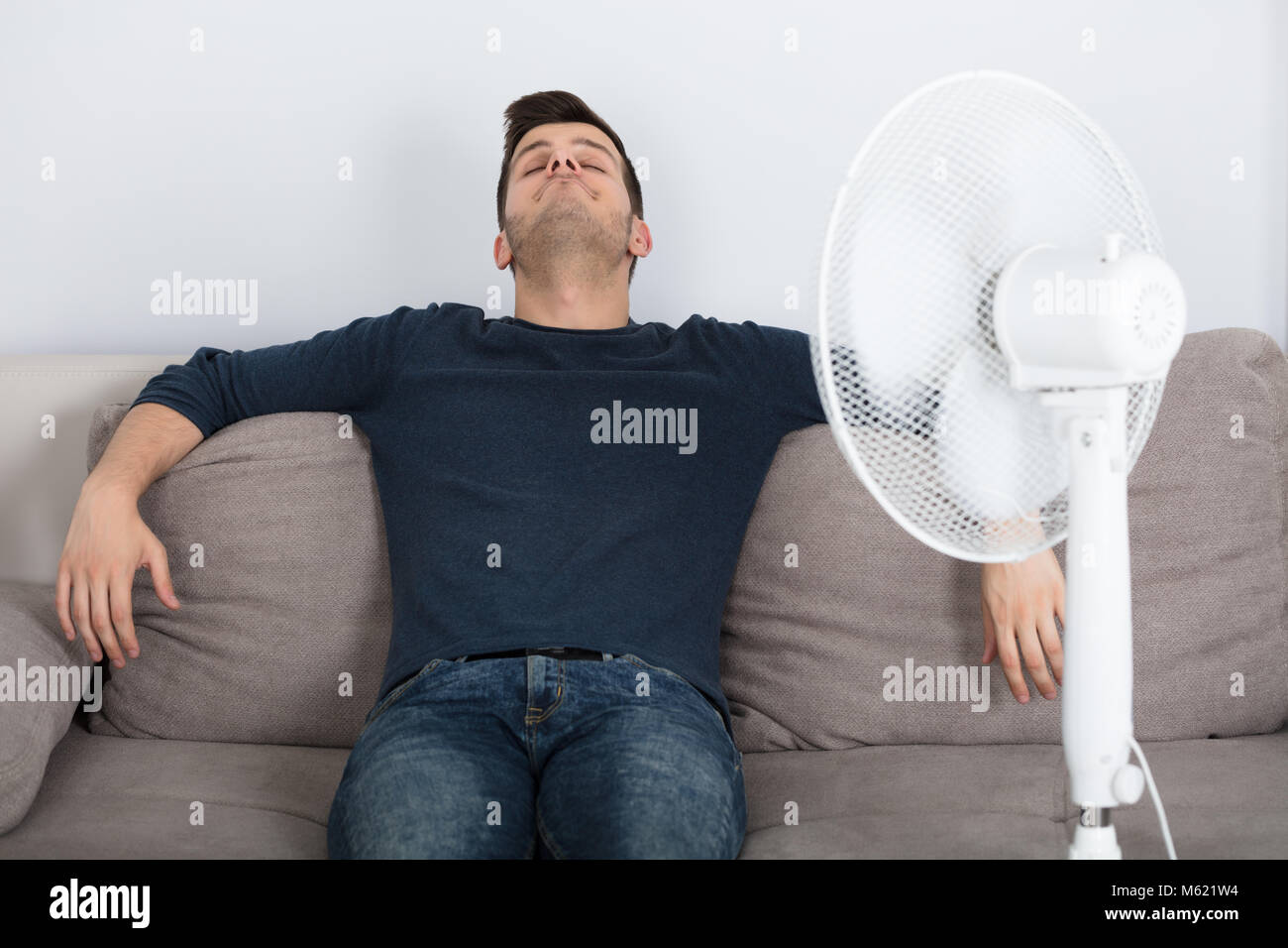 Young Man Sitting On Couch Cooling Off With Fan During Hot Weather At ...