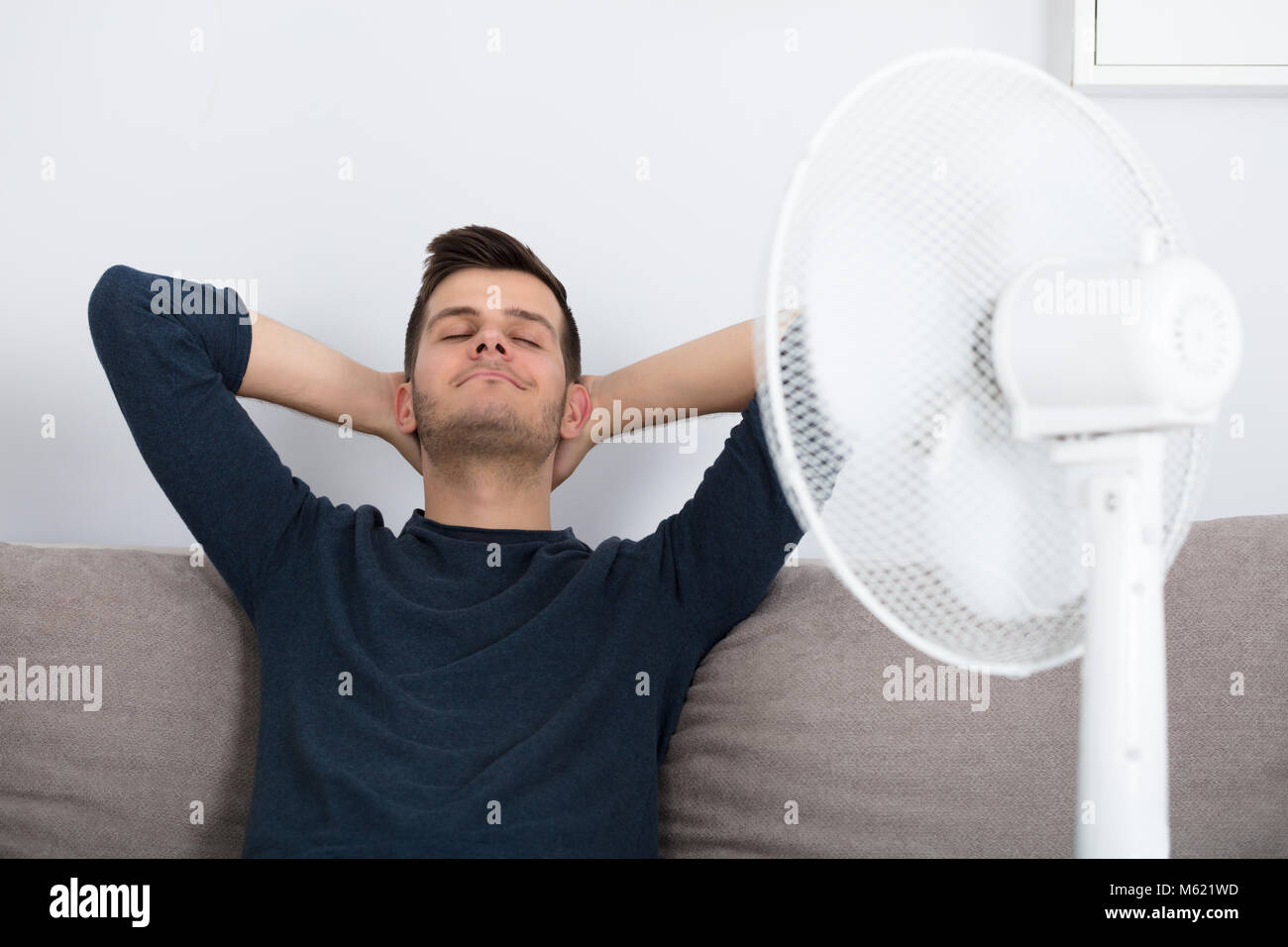 Young Man Sitting On Couch Cooling Off With Fan During Hot Weather At ...