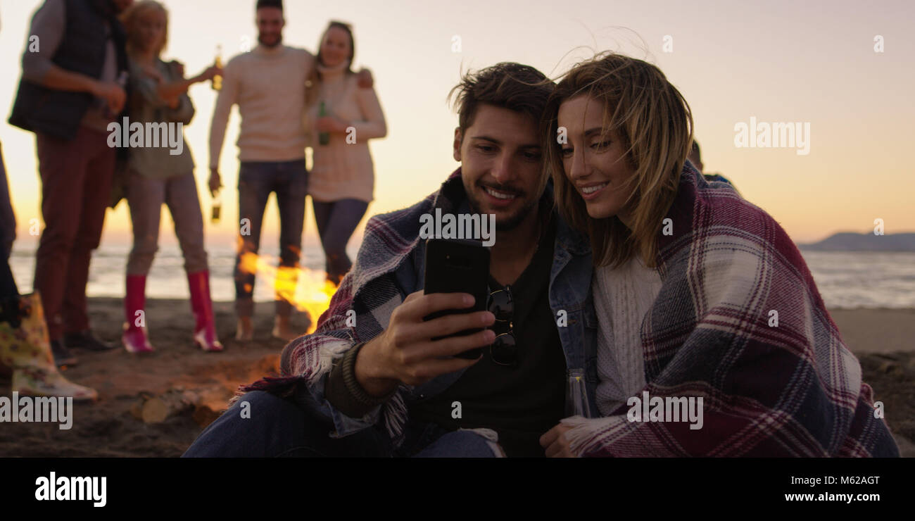 Couple using cell phone during beach party with friends drinking beer ...