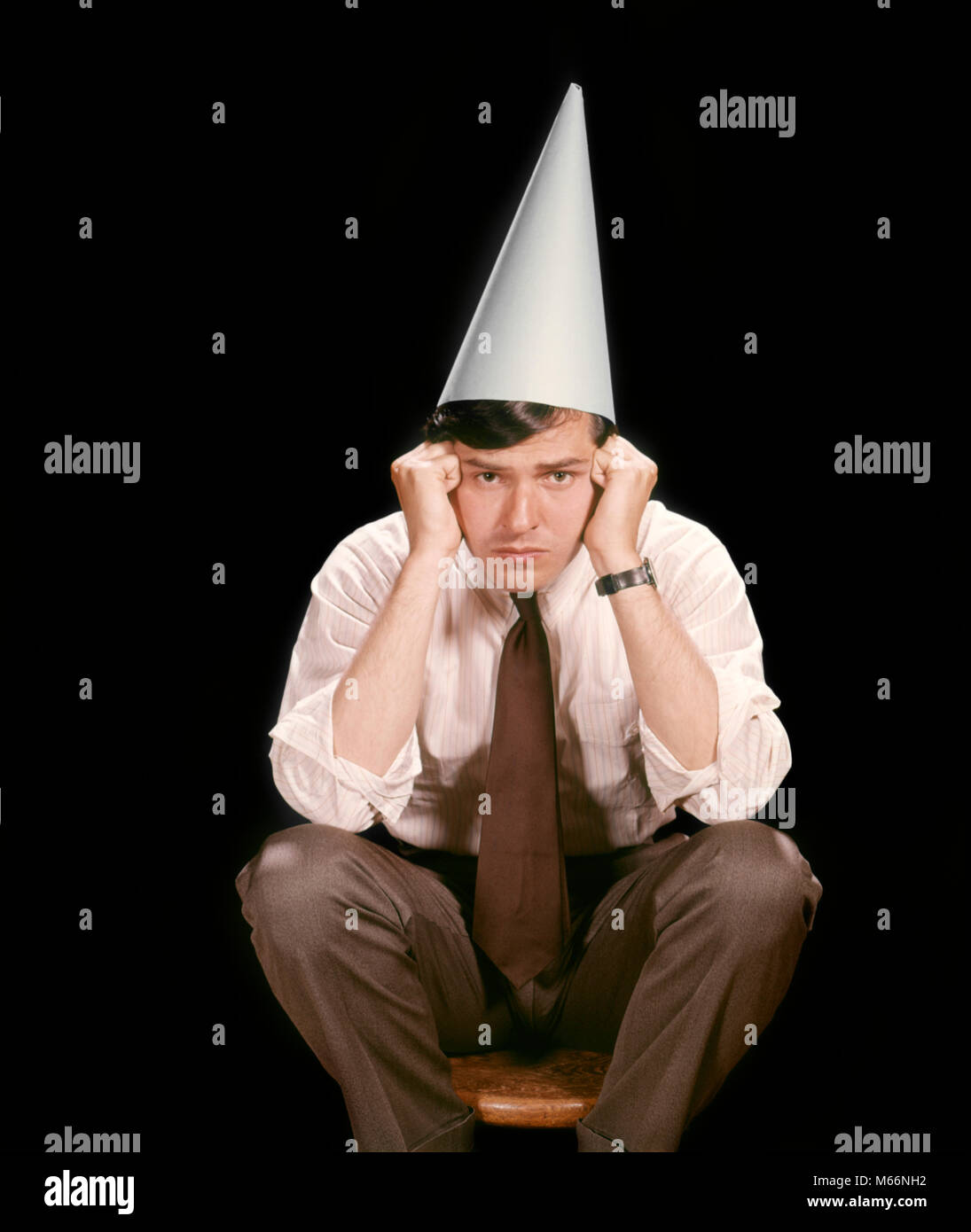 1960s UNHAPPY YOUNG MAN SITTING ON STOOL WEARING DUNCE CAP LOOKING AT ...