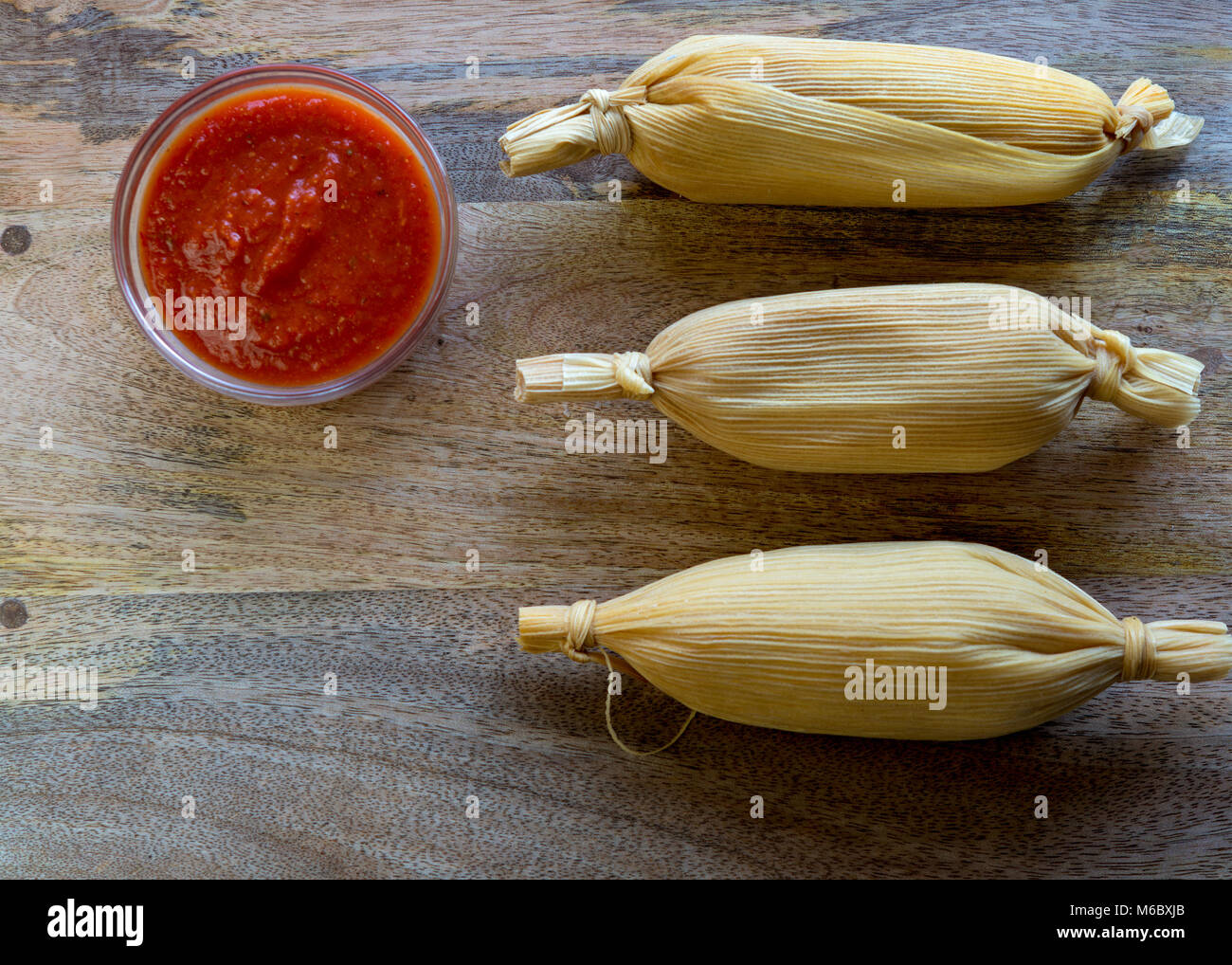 Three whole, wrapped tamales on wooden table served with hot tomato ...