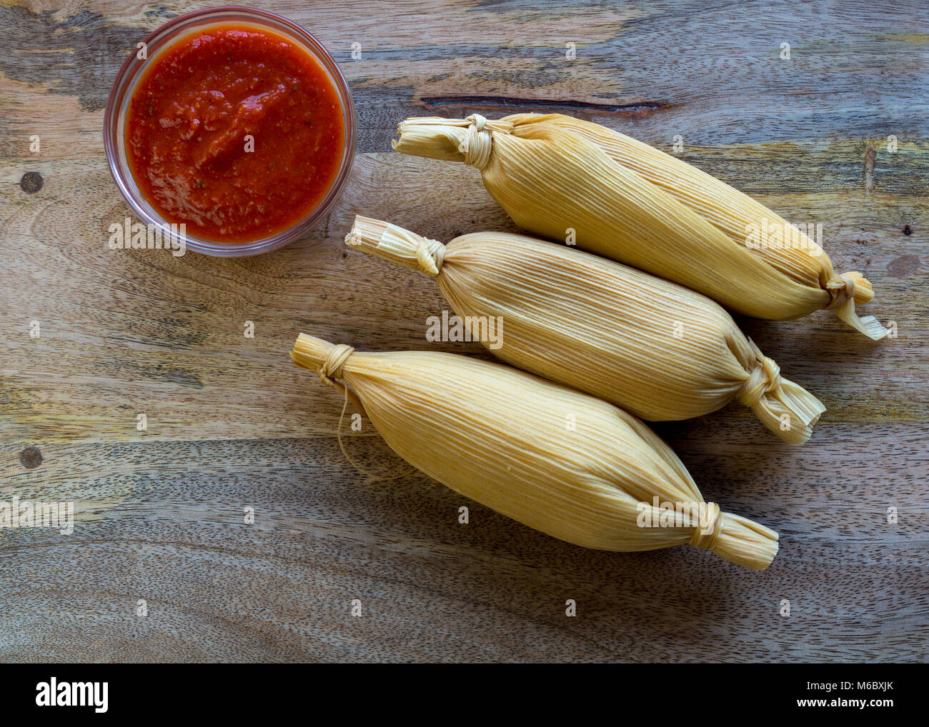 Three whole, wrapped tamales on wooden table served with hot tomato ...