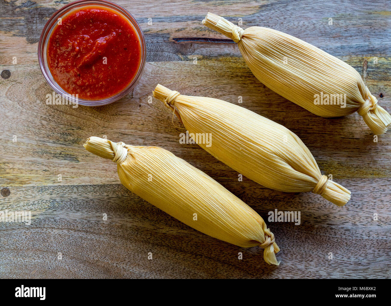 Three whole, wrapped tamales on wooden table served with hot tomato ...