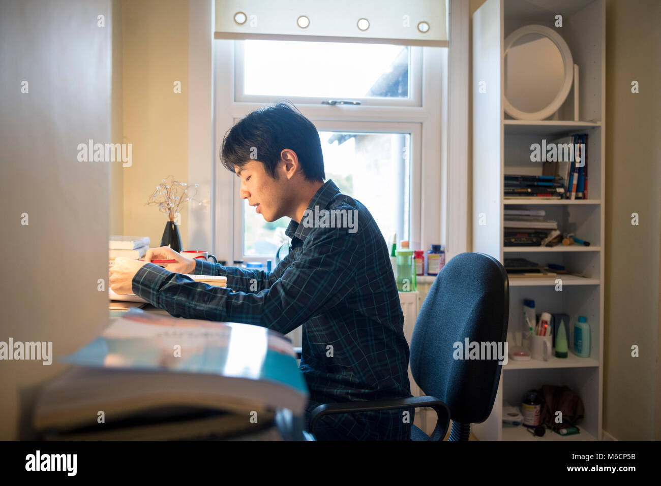 a young asian student works in his bedroom reading a book and revising ...