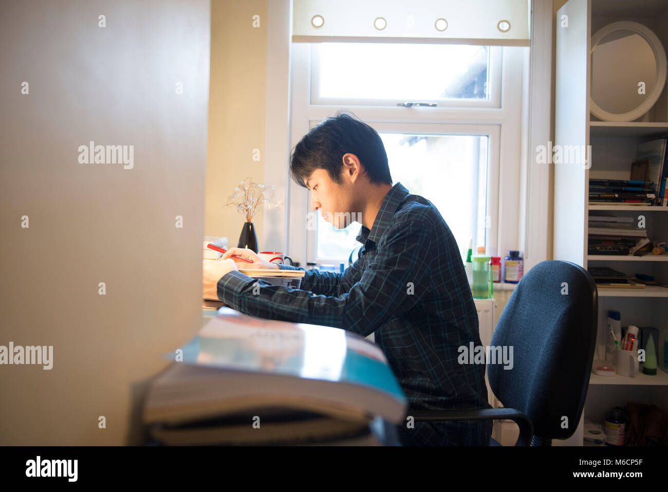 a young asian student works in his bedroom reading a book and revising ...