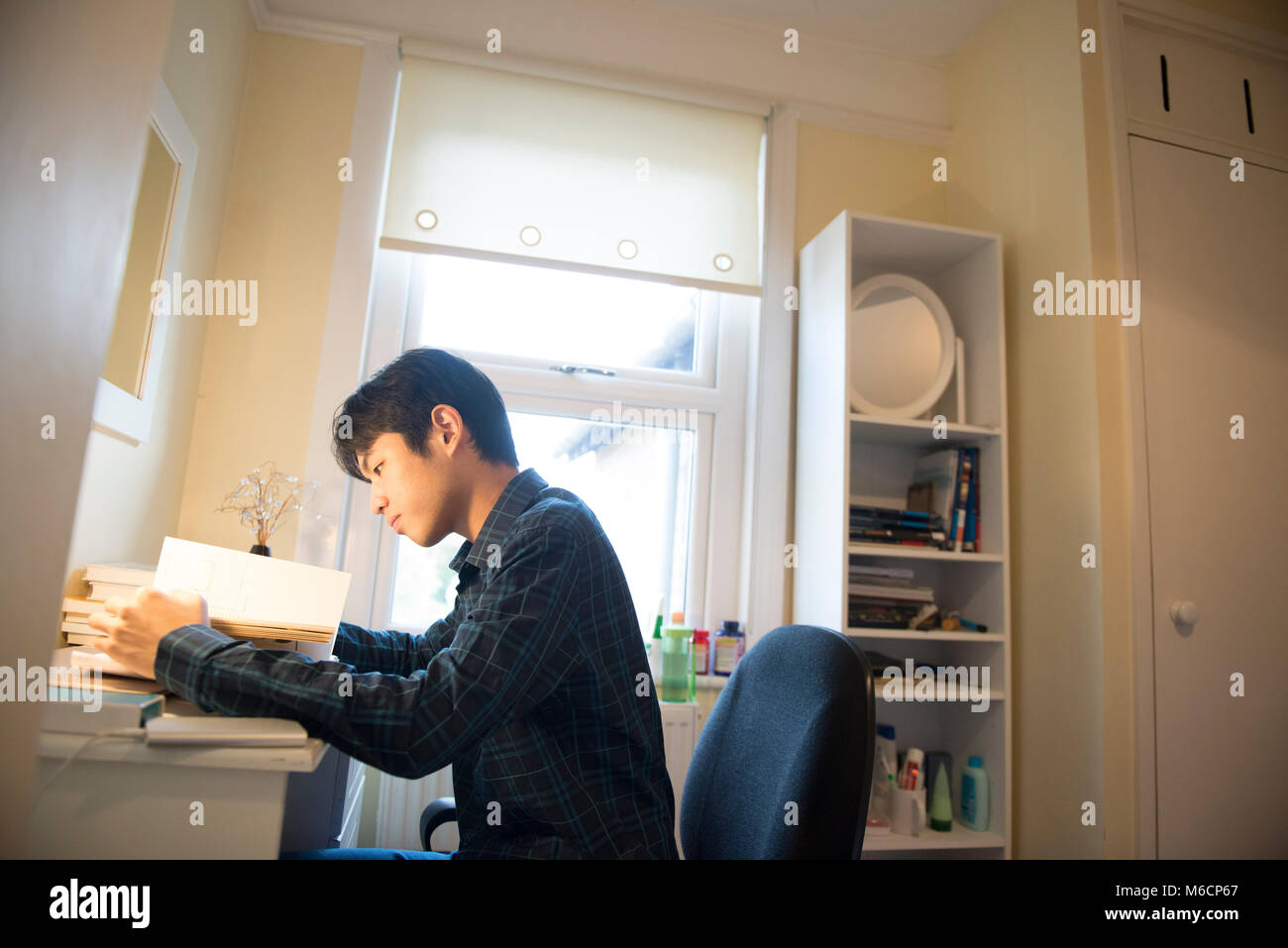 a young asian student works in his bedroom reading a book and revising ...
