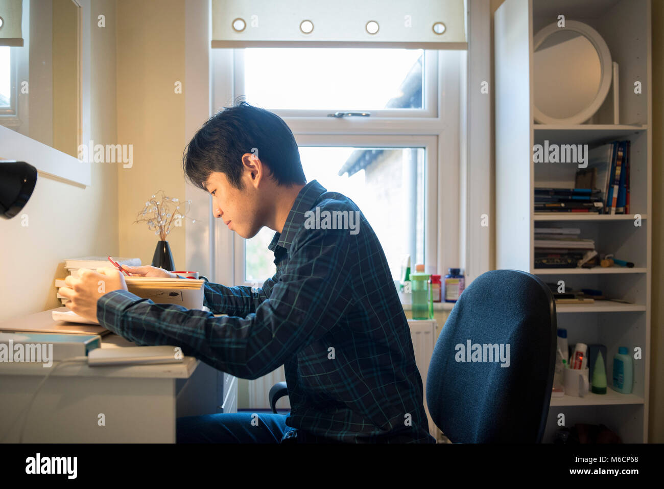 a young asian student works in his bedroom reading a book and revising ...