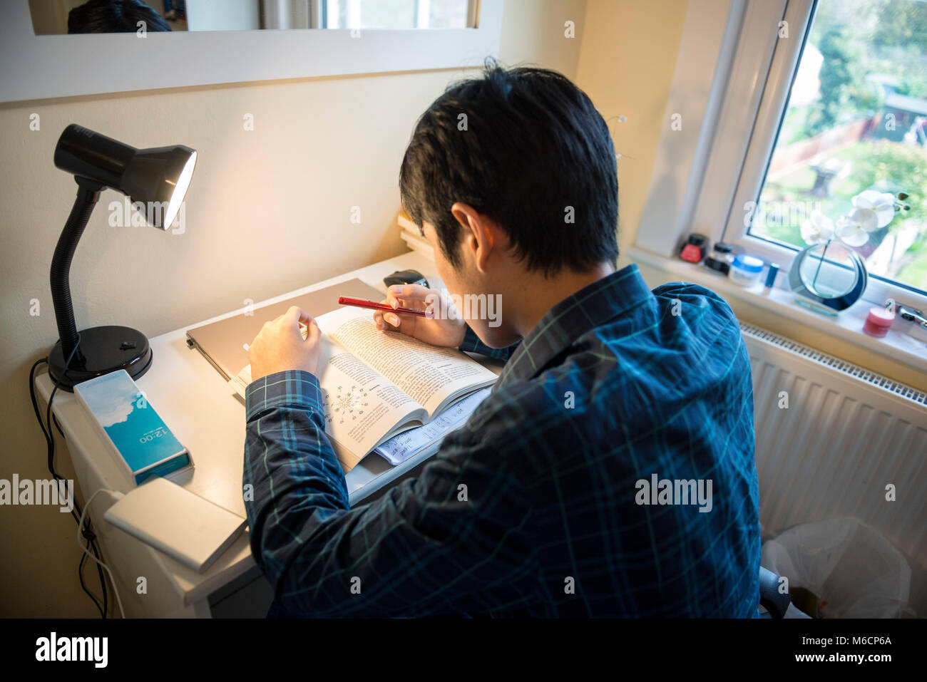 a young asian student works in his bedroom reading a book and revising ...