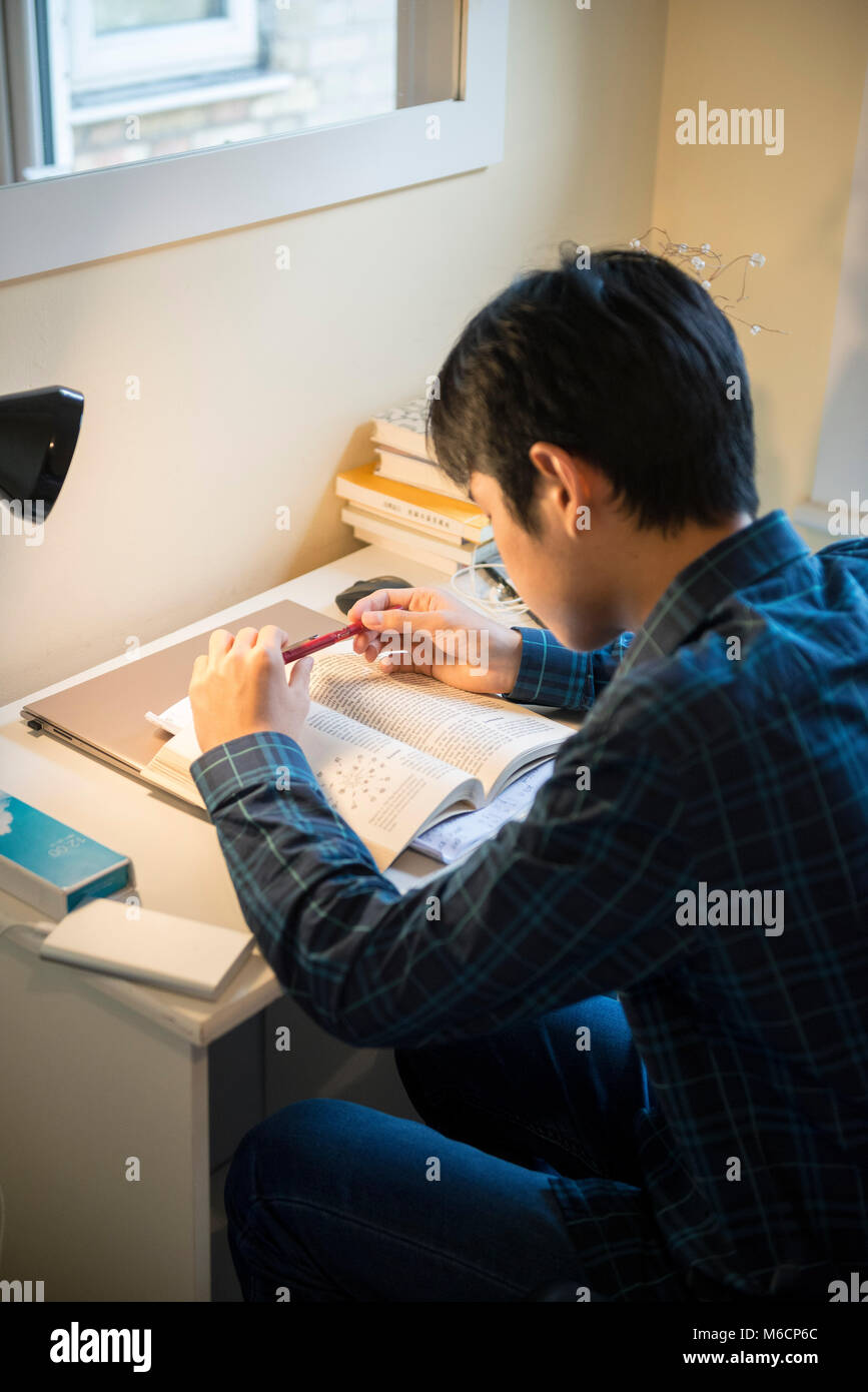 a young asian student works in his bedroom reading a book and revising ...