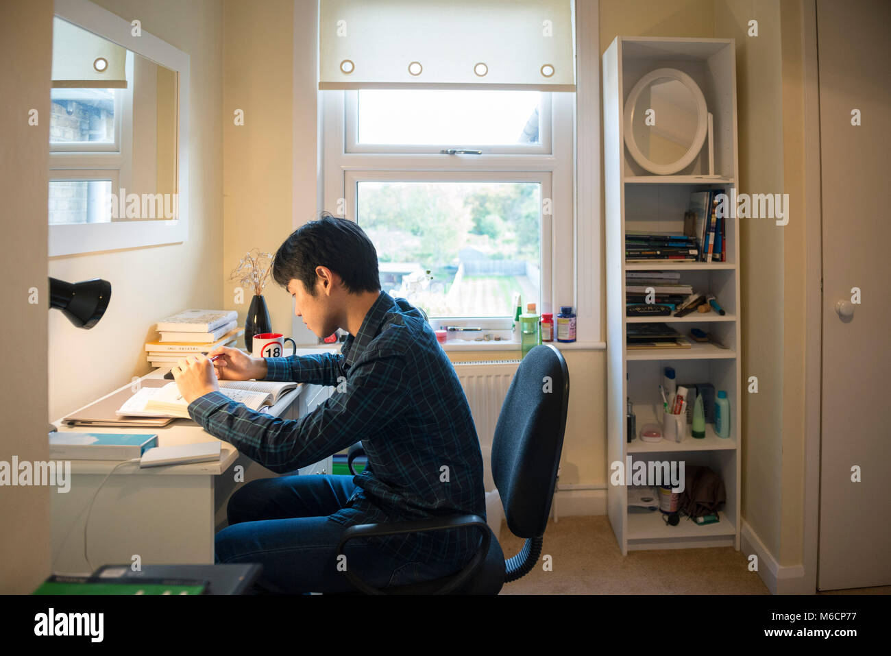 a young asian student works in his bedroom reading a book and revising ...
