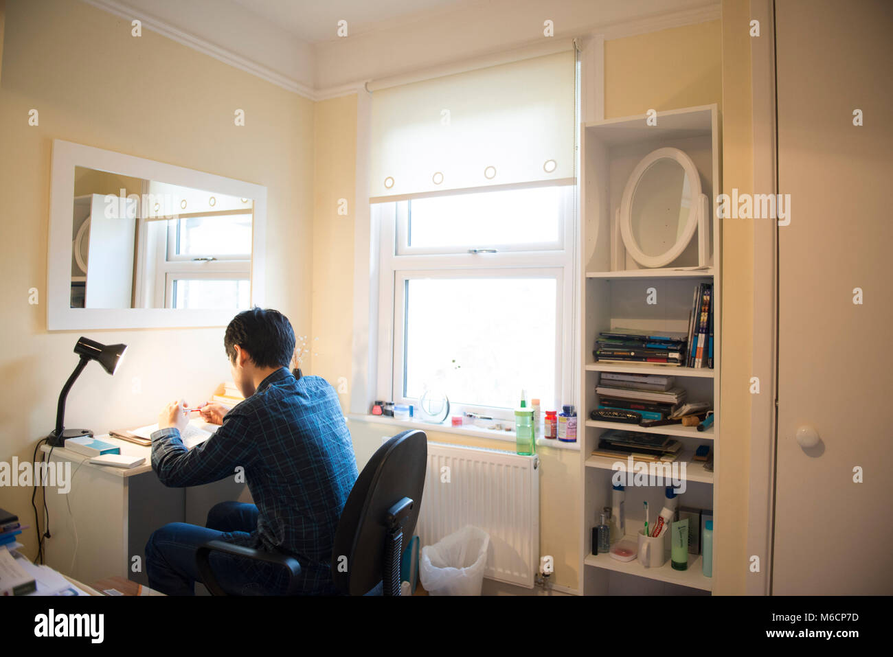 a young asian student works in his bedroom reading a book and revising ...