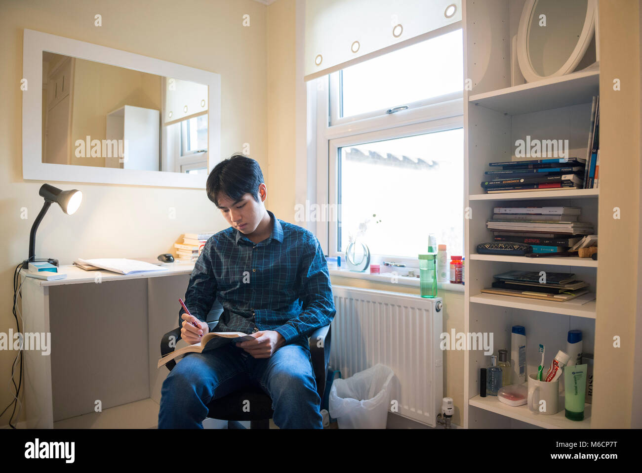 a young asian student works in his bedroom reading a book and revising ...