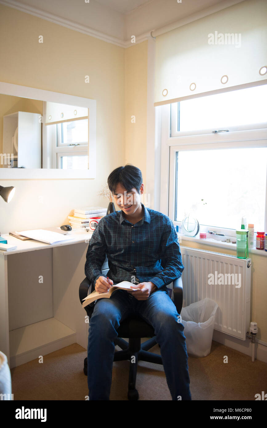 a young asian student works in his bedroom reading a book and revising ...