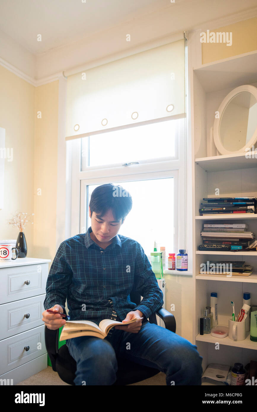 a young asian student works in his bedroom reading a book and revising ...