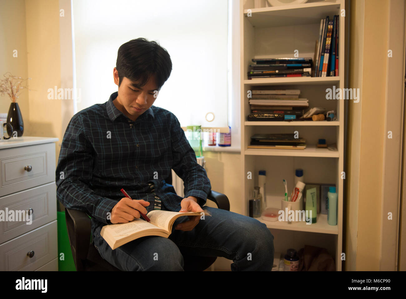 a young asian student works in his bedroom reading a book and revising ...