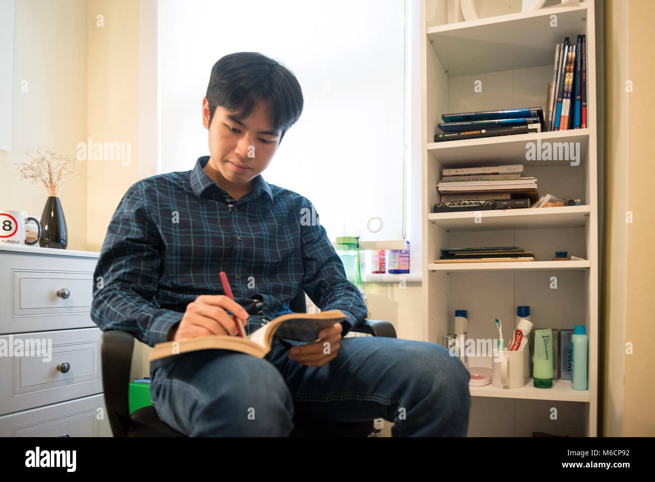 a young asian student works in his bedroom reading a book and revising ...
