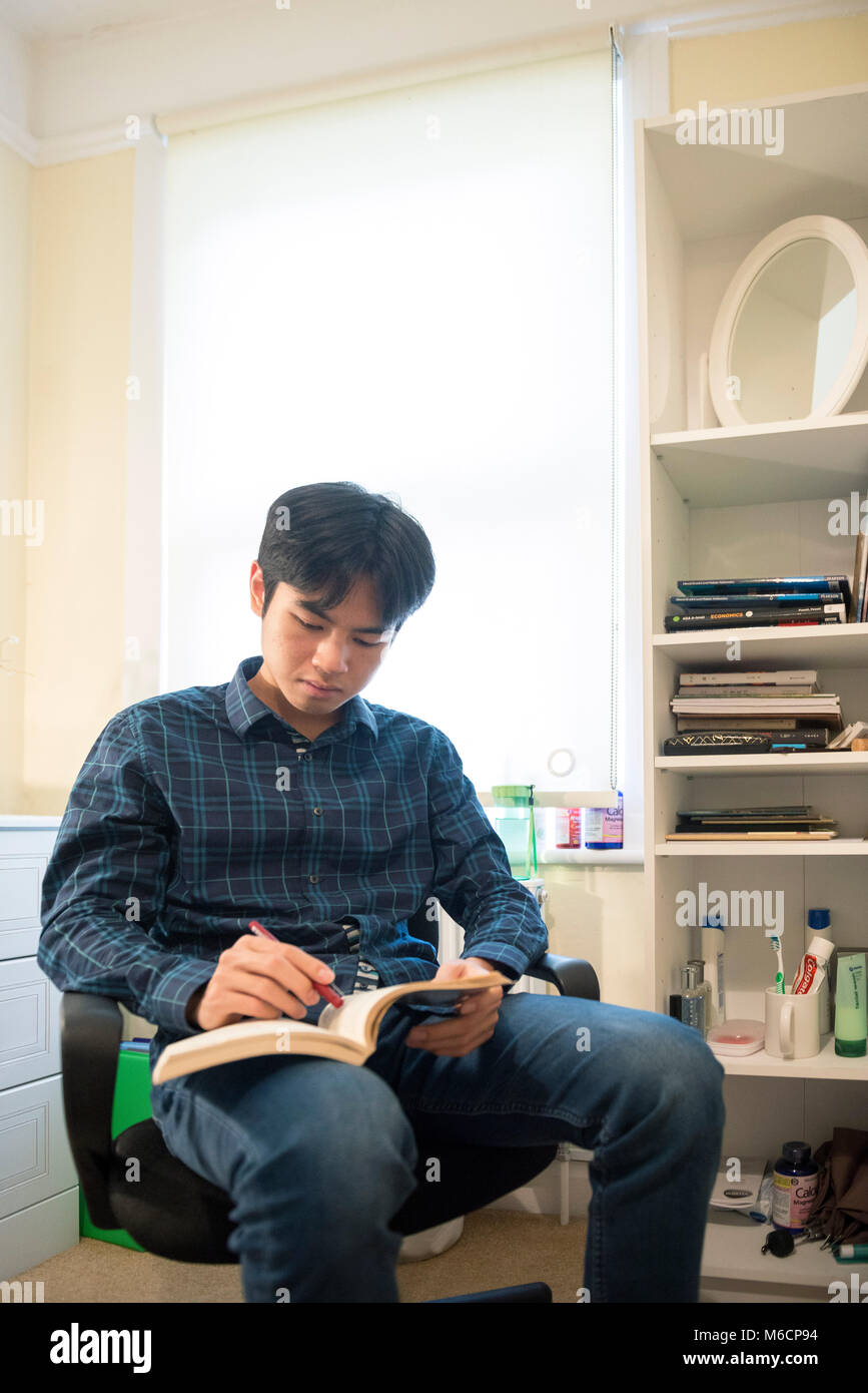 a young asian student works in his bedroom reading a book and revising ...