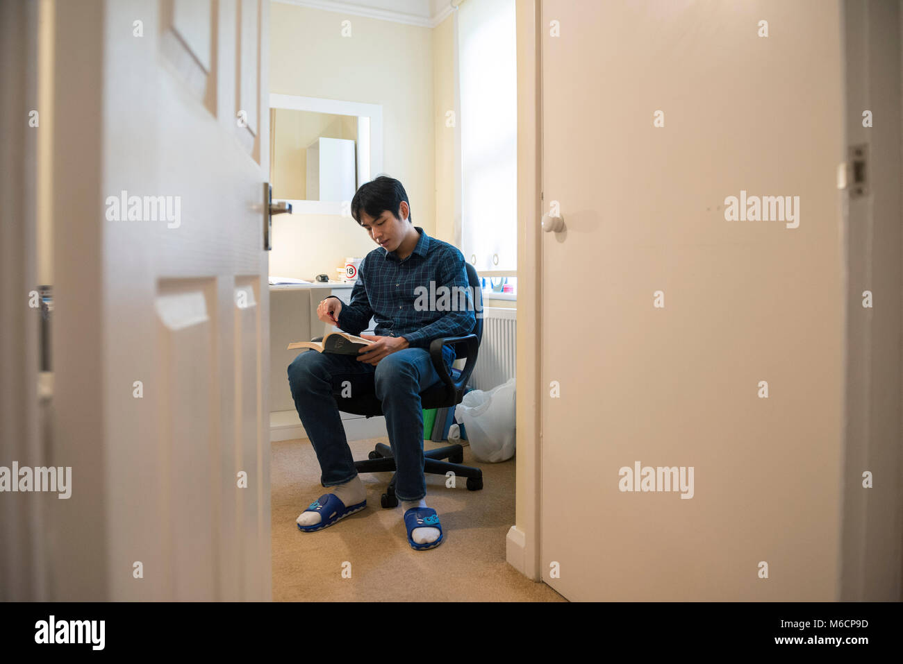a young asian student works in his bedroom reading a book and revising ...