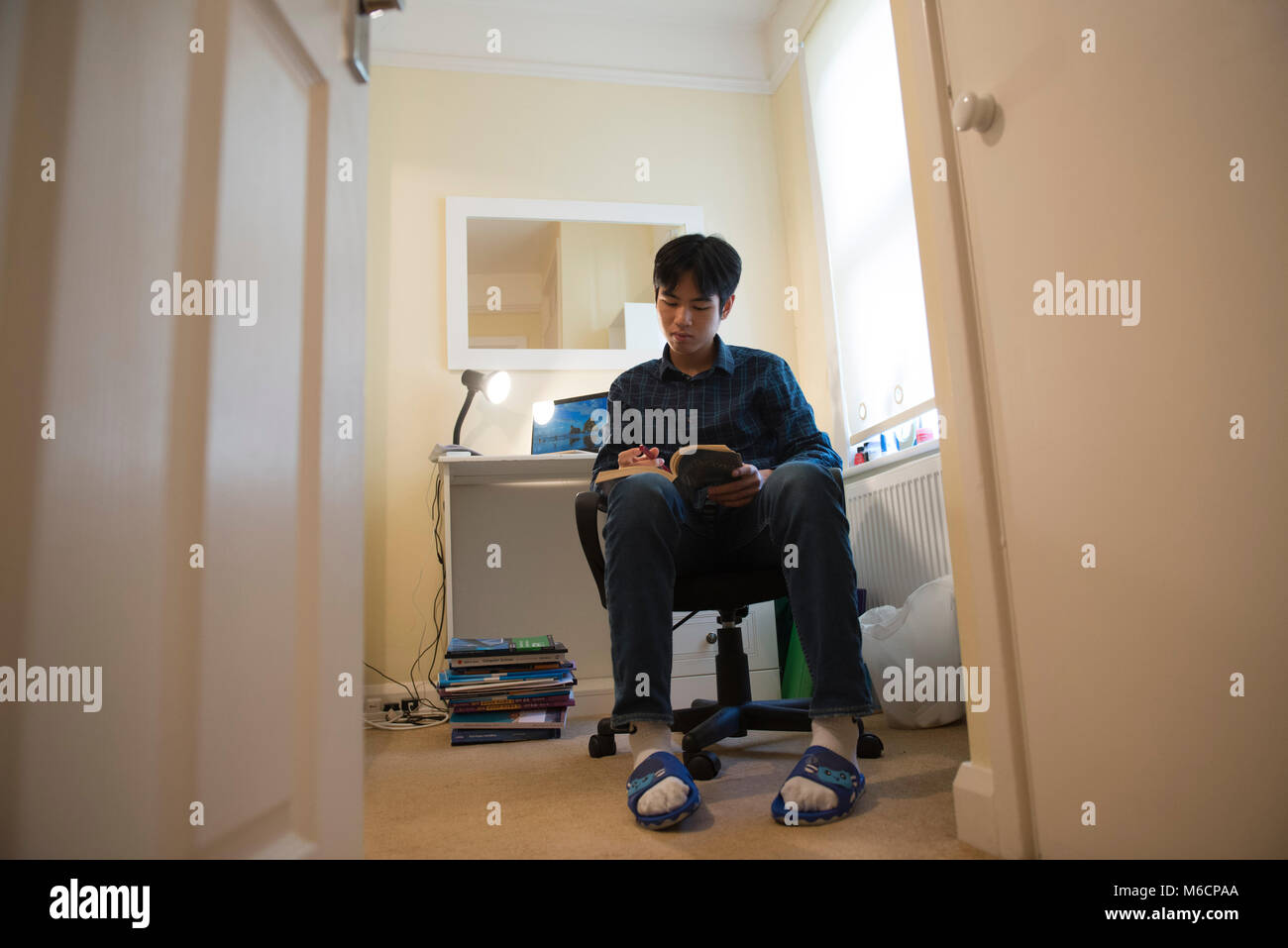 a young asian student works in his bedroom reading a book and revising ...