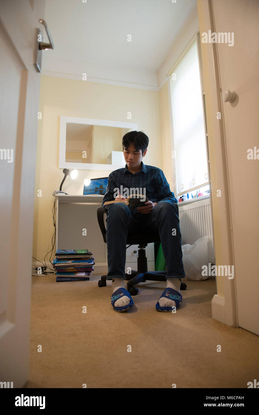 a young asian student works in his bedroom reading a book and revising ...
