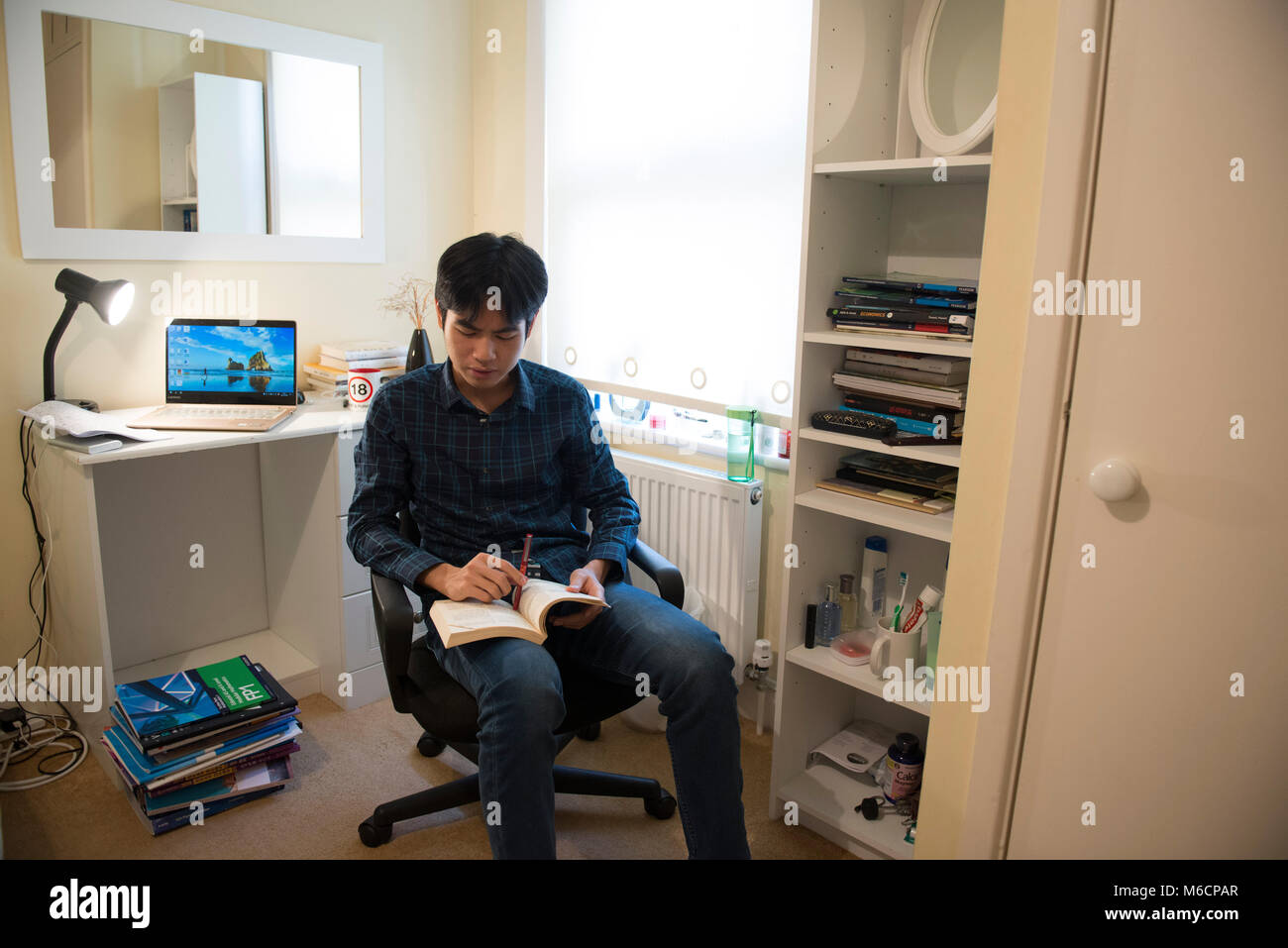 a young asian student works in his bedroom reading a book and revising ...