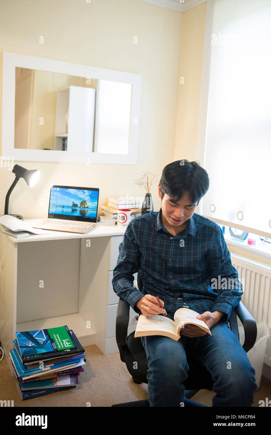 a young asian student works in his bedroom reading a book and revising ...
