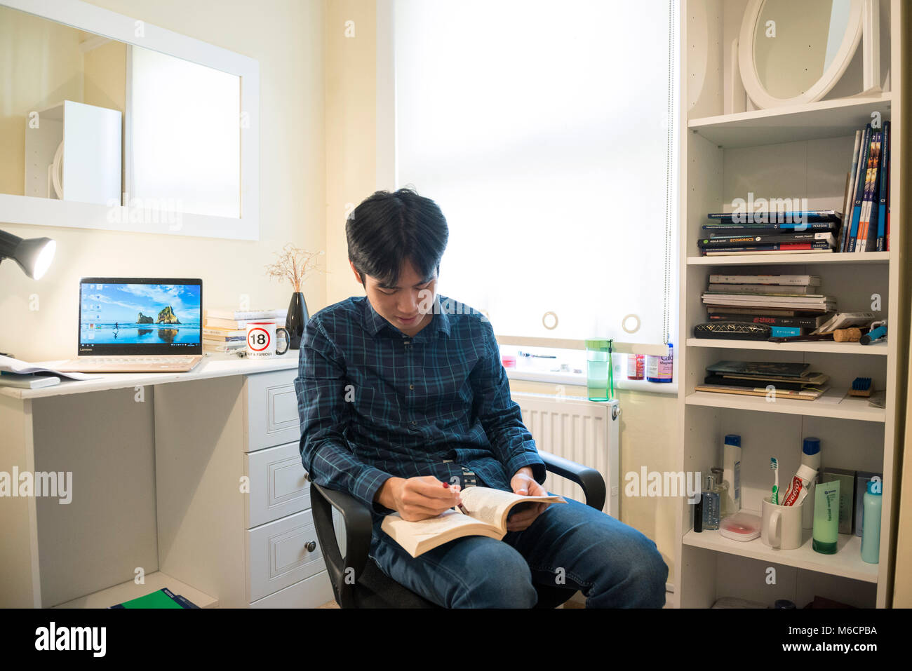 a young asian student works in his bedroom reading a book and revising ...