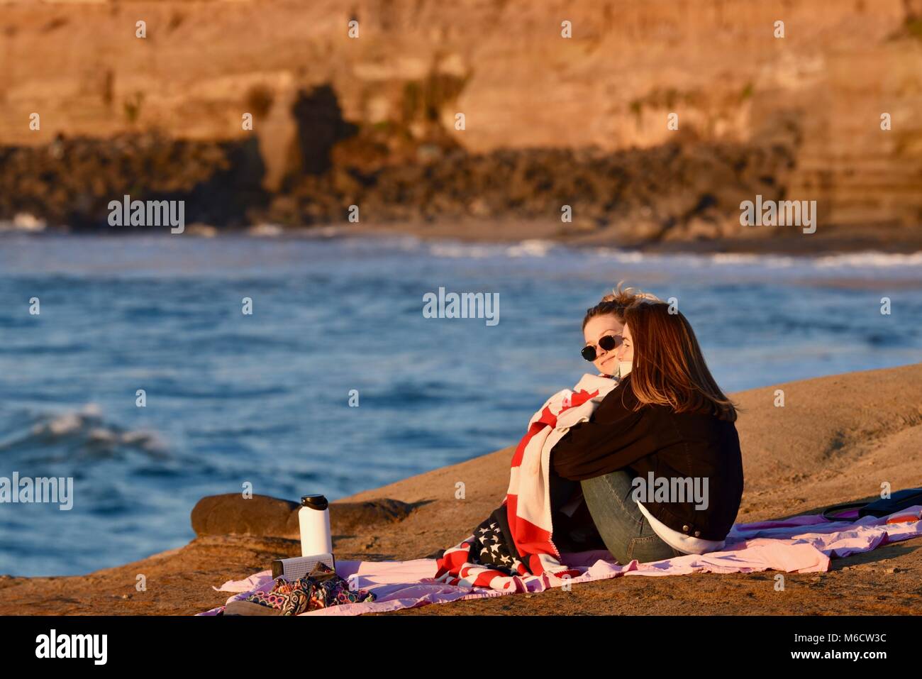 Two young women in blanket enjoy a picnic at sunset along golden cliffs ...