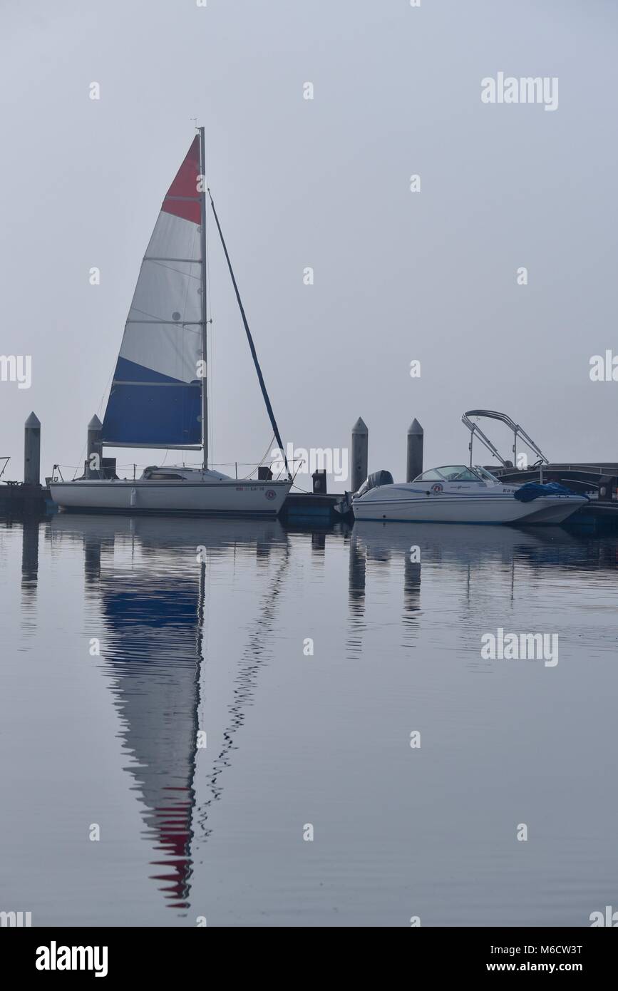Sailboat with red, white and blue main sail up, tied up at dock, with ...