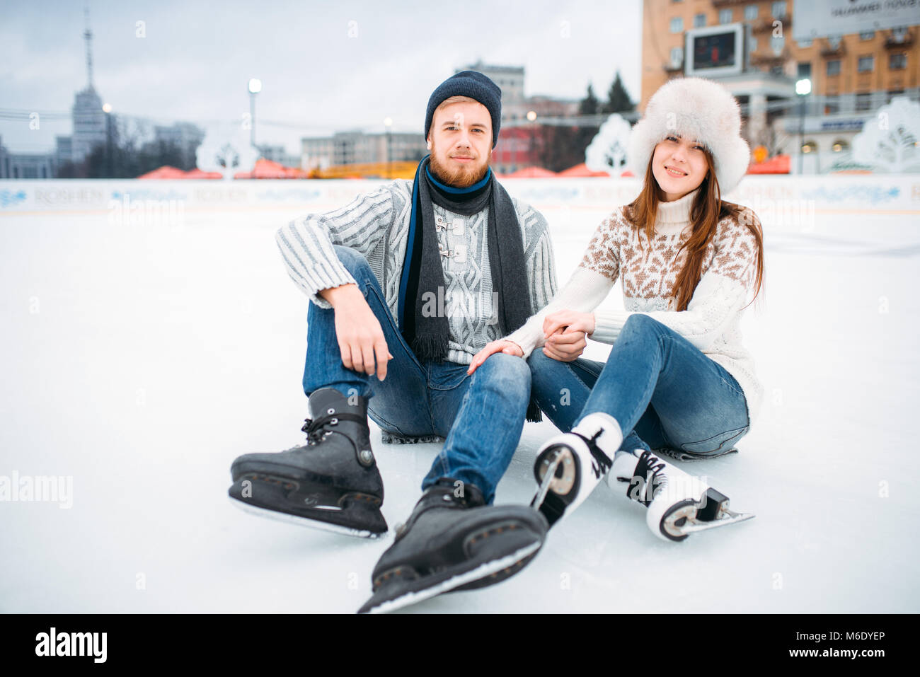 Young love couple in skates sitting on ice, skating rink. Winter ice ...