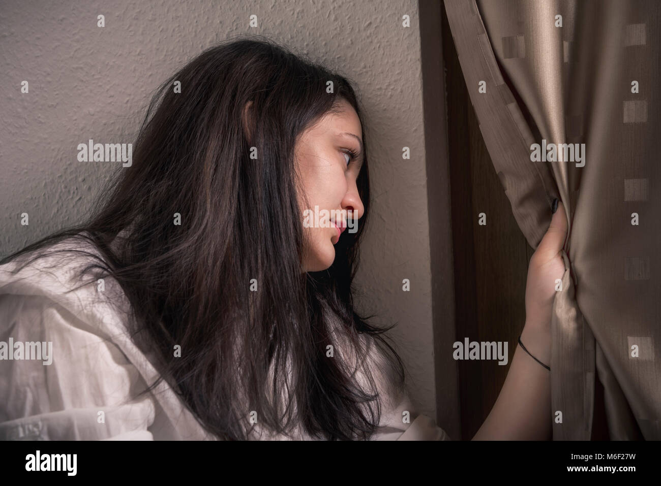 Young brunette woman peeking through the window, from behind curtains ...