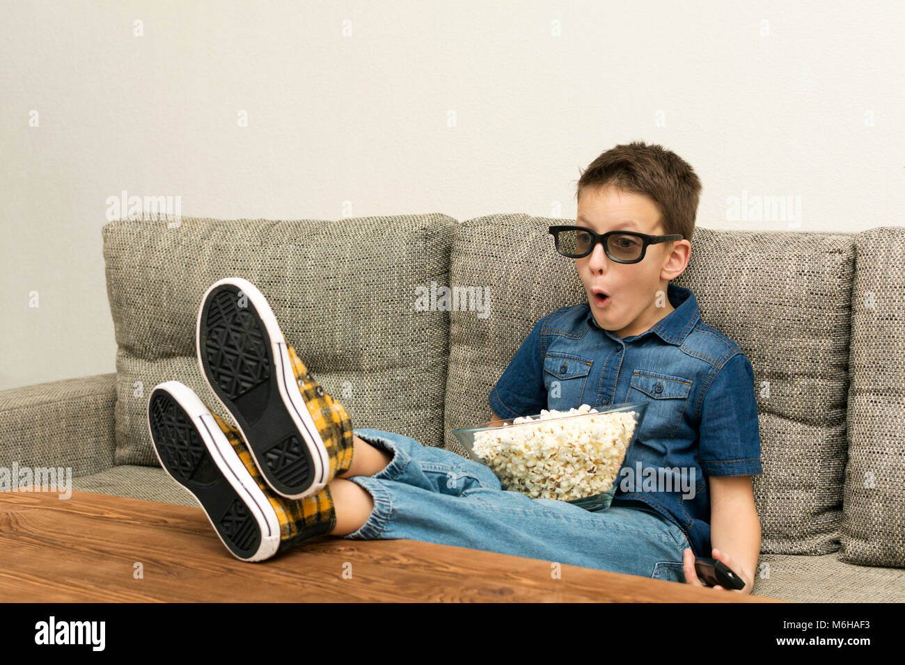 Small boy on the couch watching TV and eating popcorn. Legs on table ...