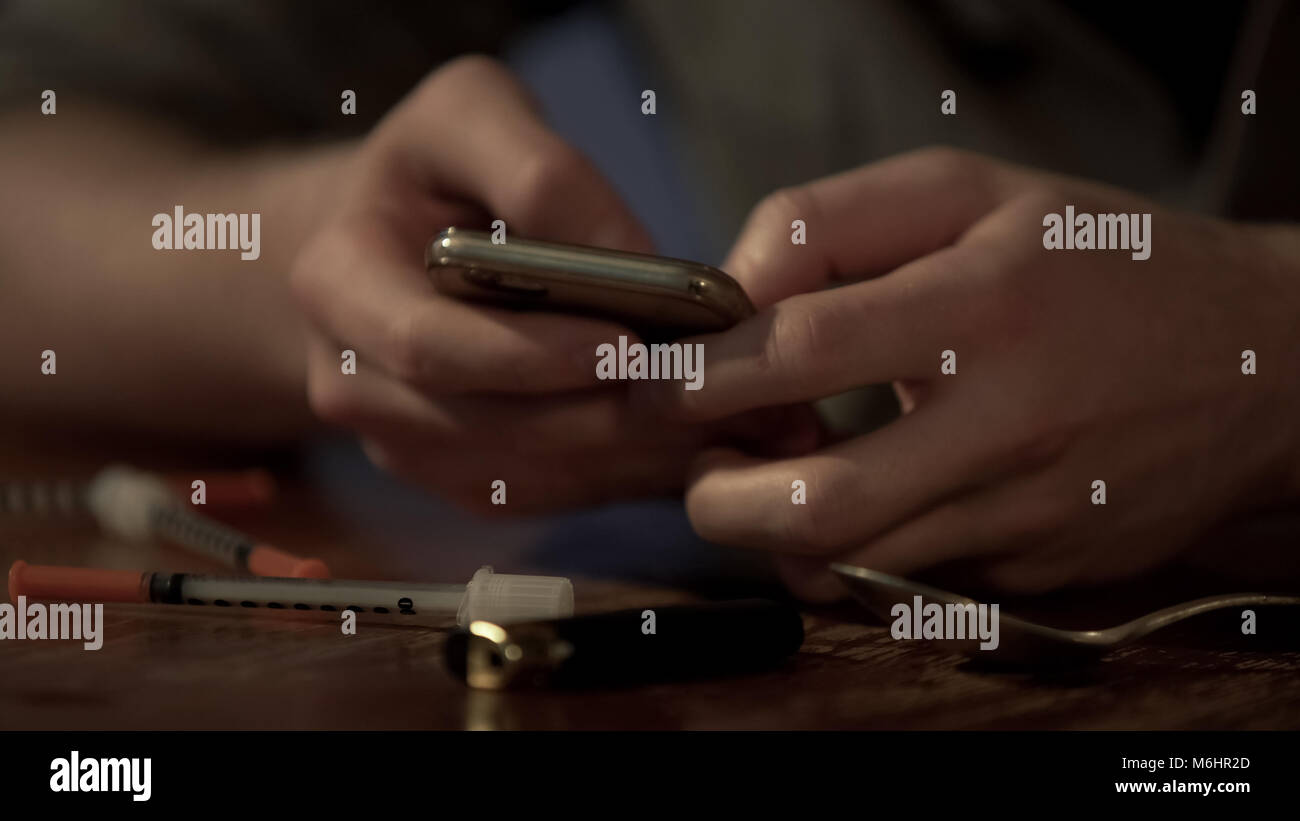 Male junkie typing message on smartphone, many syringes lie on table ...